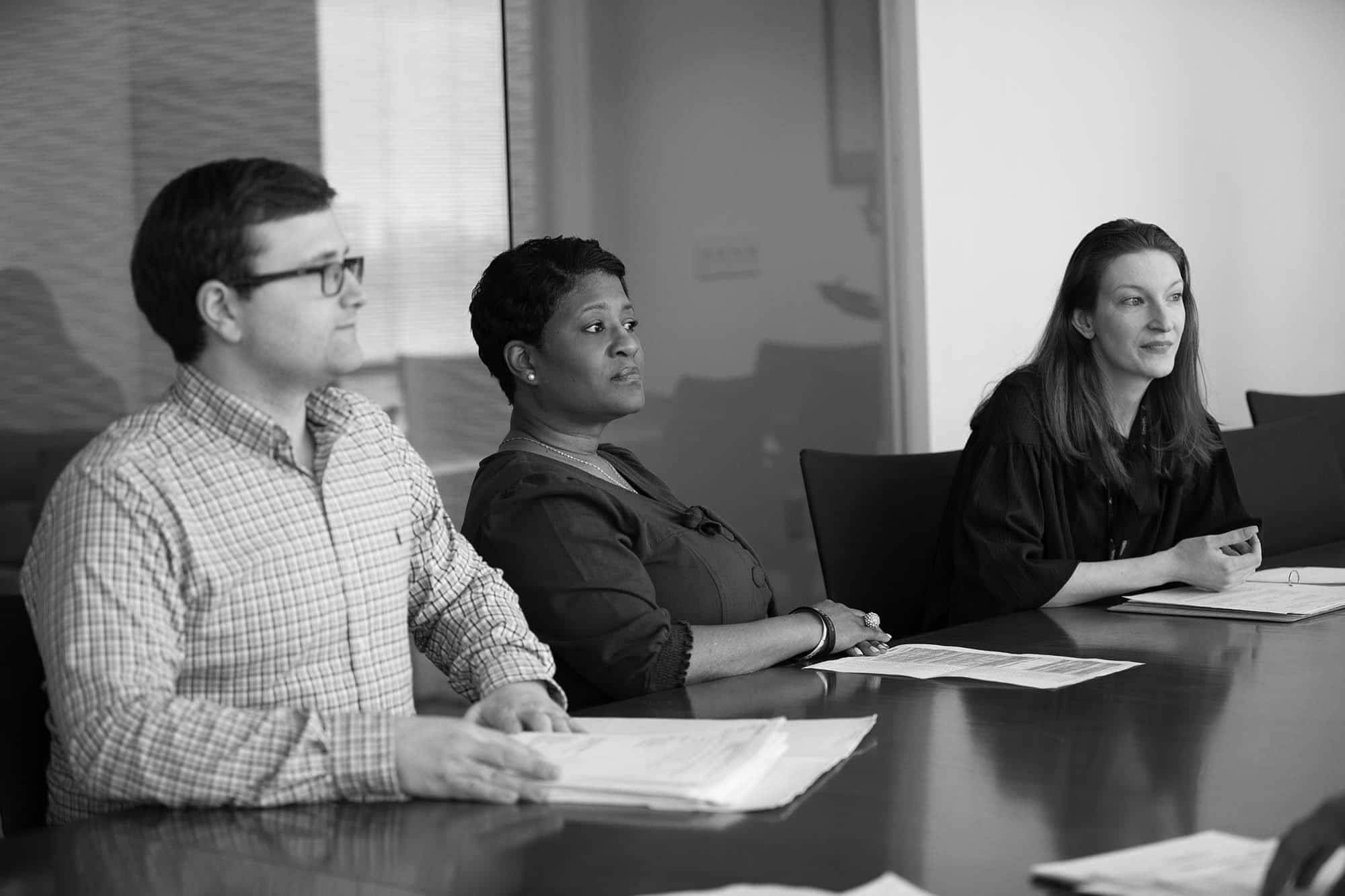 Three people sit at a conference table with papers in front of them ...